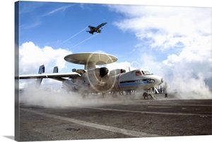 An F/A-18C Hornet flies overhead as an E-2C Hawkeye prepares to launch ...